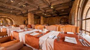 Um restaurante com paredes de tijolo, vigas de madeira e mesas dispostas com toalhas brancas e jogos americanos de veludo laranja.