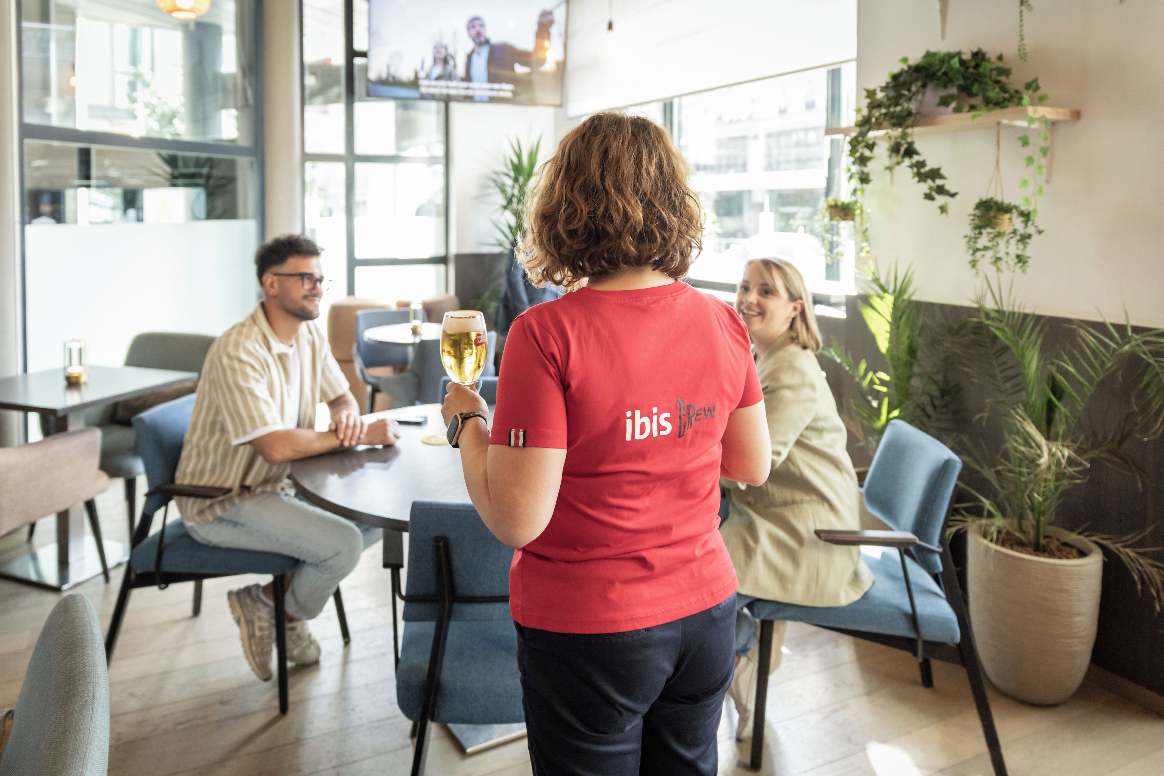 Pessoa vestindo uma camisa vermelha com a inscrição 'ibis Brew' nas costas, em um quarto com uma mesa, cadeiras e janelas amplas.
