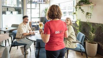 Pessoa vestindo uma camisa vermelha com a inscrição 'ibis Brew' nas costas, em um quarto com uma mesa, cadeiras e janelas amplas.
