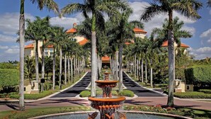 Um jardim simétrico com uma fonte central, palmeiras e um edifício com telhados vermelhos ao fundo.