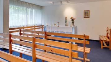 Interior de igreja com bancos de madeira, altar branco com objetos religiosos e uma grande janela com cortinas leves.
