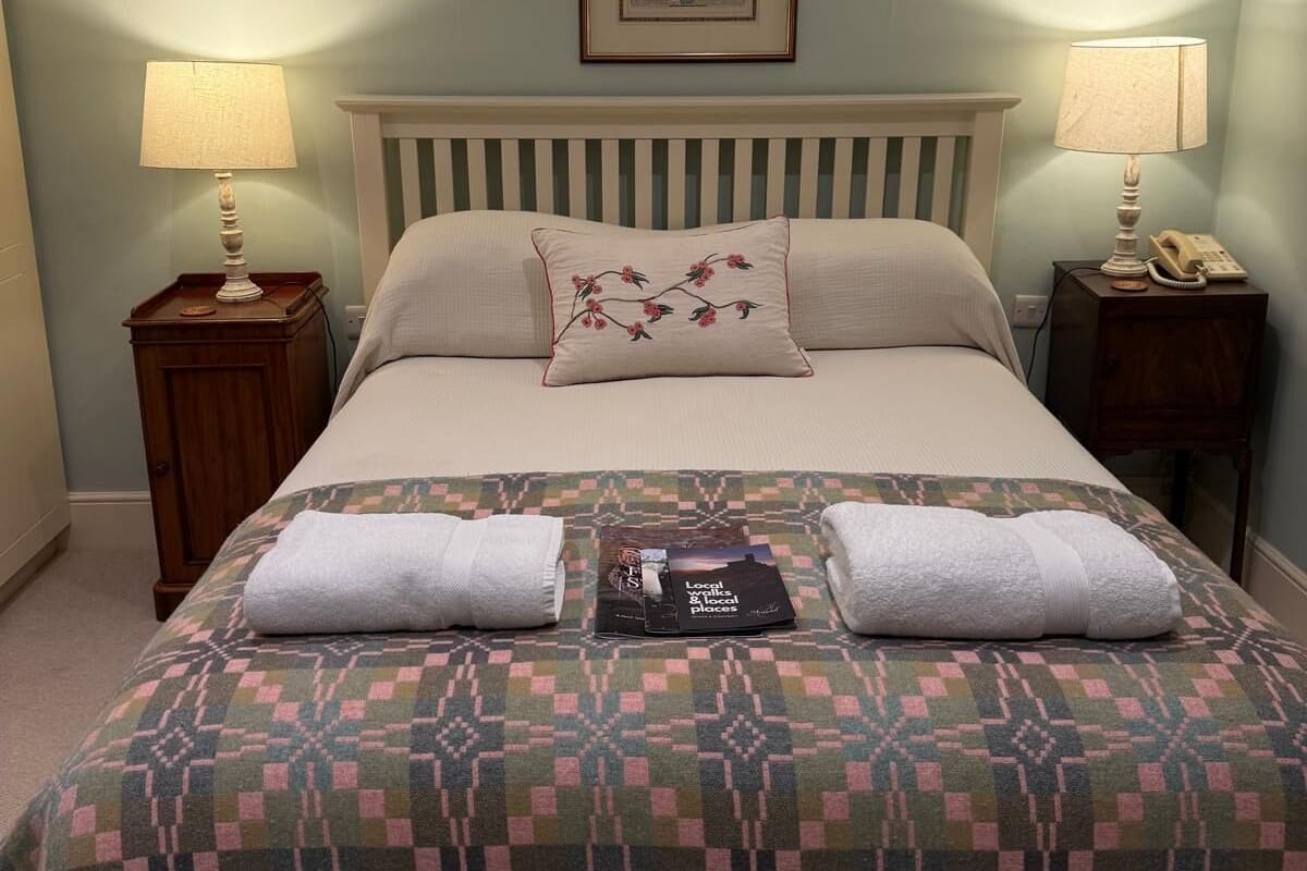Cama bem arrumada com colcha estampada, duas luminárias laterais, um quadro emoldurado acima da cabeceira e um folheto sobre a cama.
