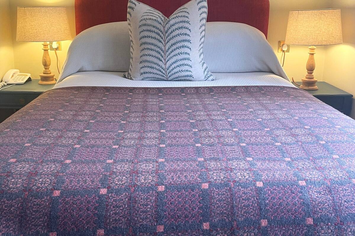 Uma cama bem arrumada com colcha estampada, duas almofadas com desenho de folhas, cabeceira vermelha e um quadro na parede.
