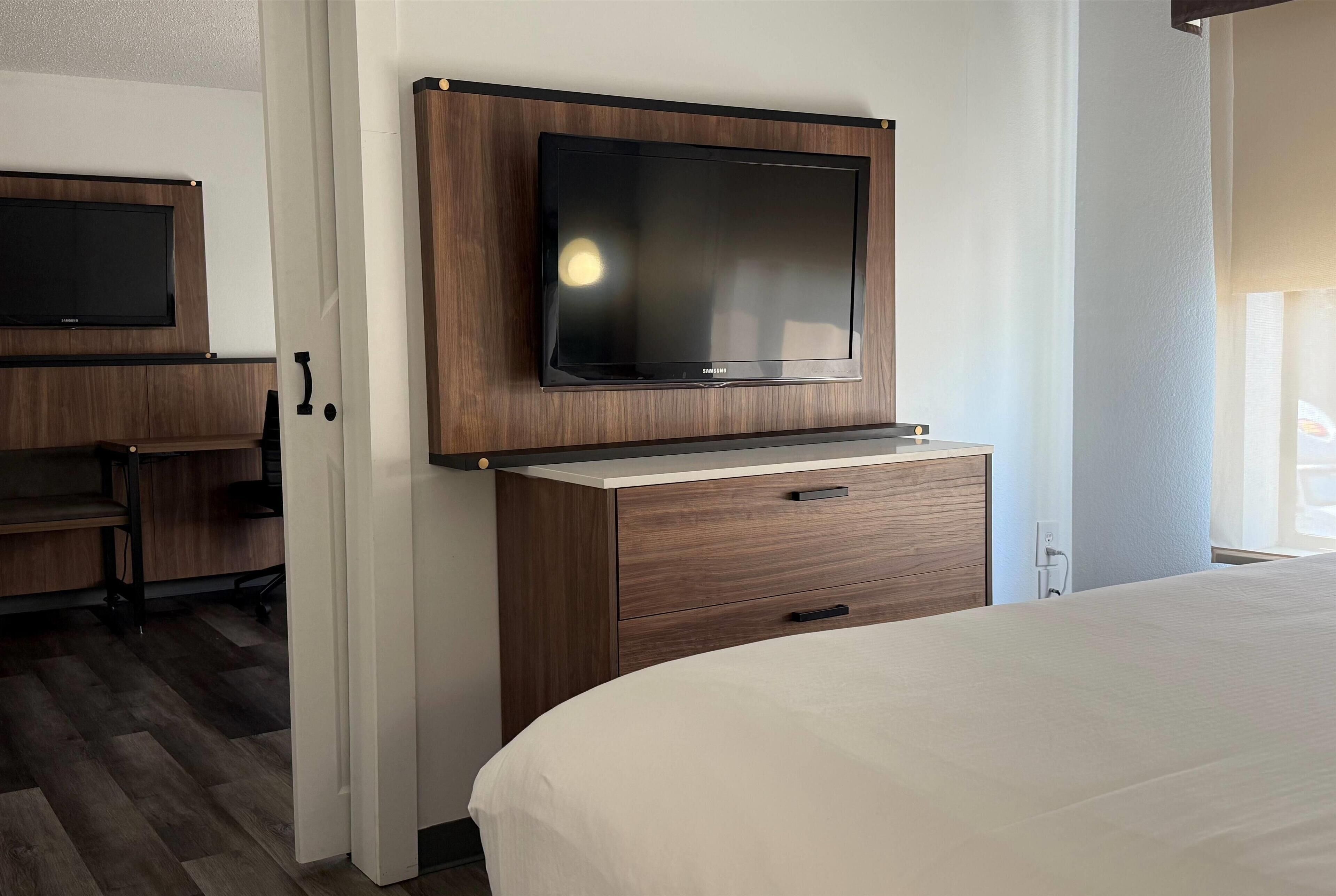 Quarto de hotel com uma televisão de tela plana grande presa a um armário de madeira, uma cama com colcha branca e uma escrivaninha com cadeira.