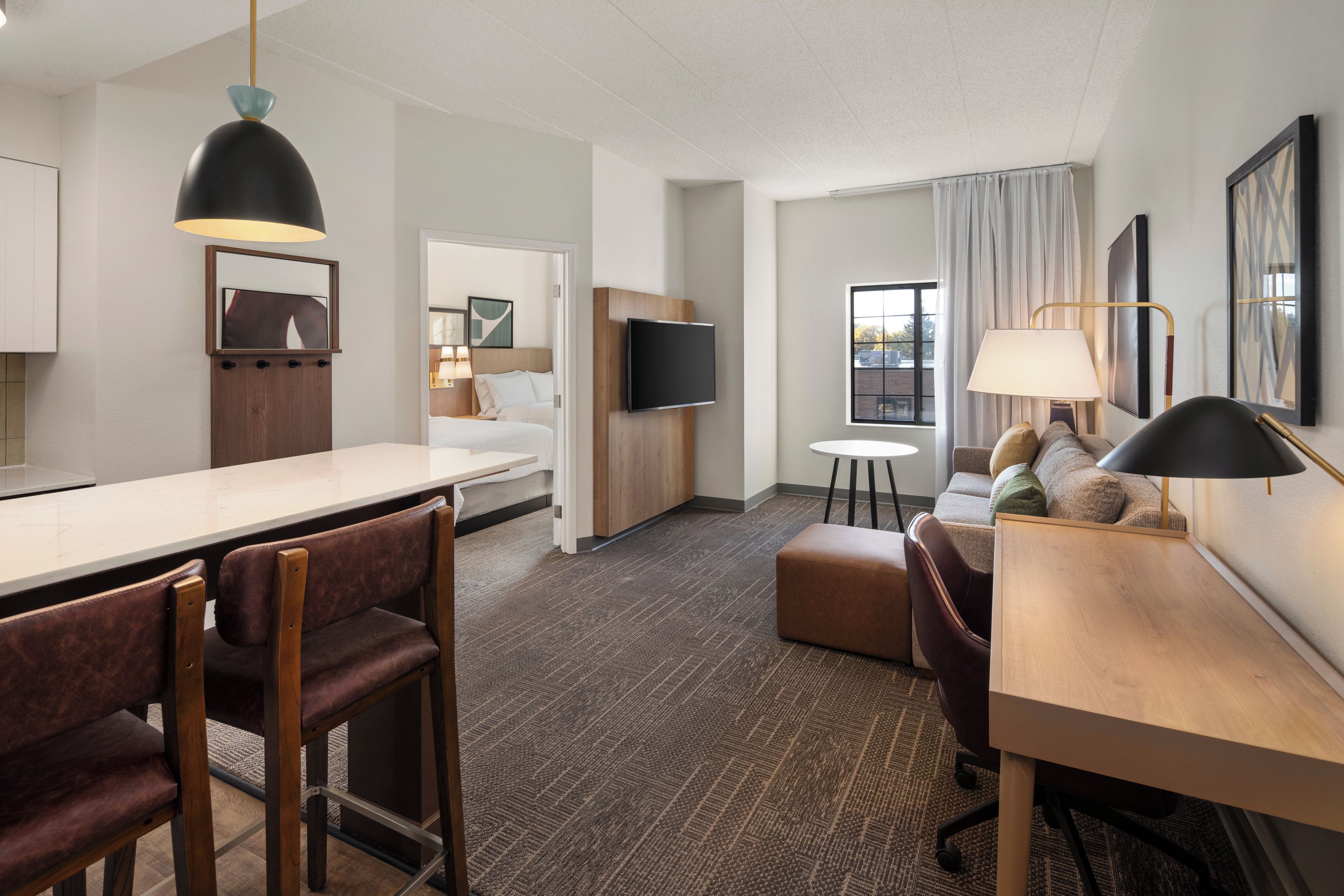 Quarto de hotel com cozinha compacta, espaço de refeições com banquetas, área de estar com sofá e poltronas, TV de tela plana na parede e janela com cortinas.