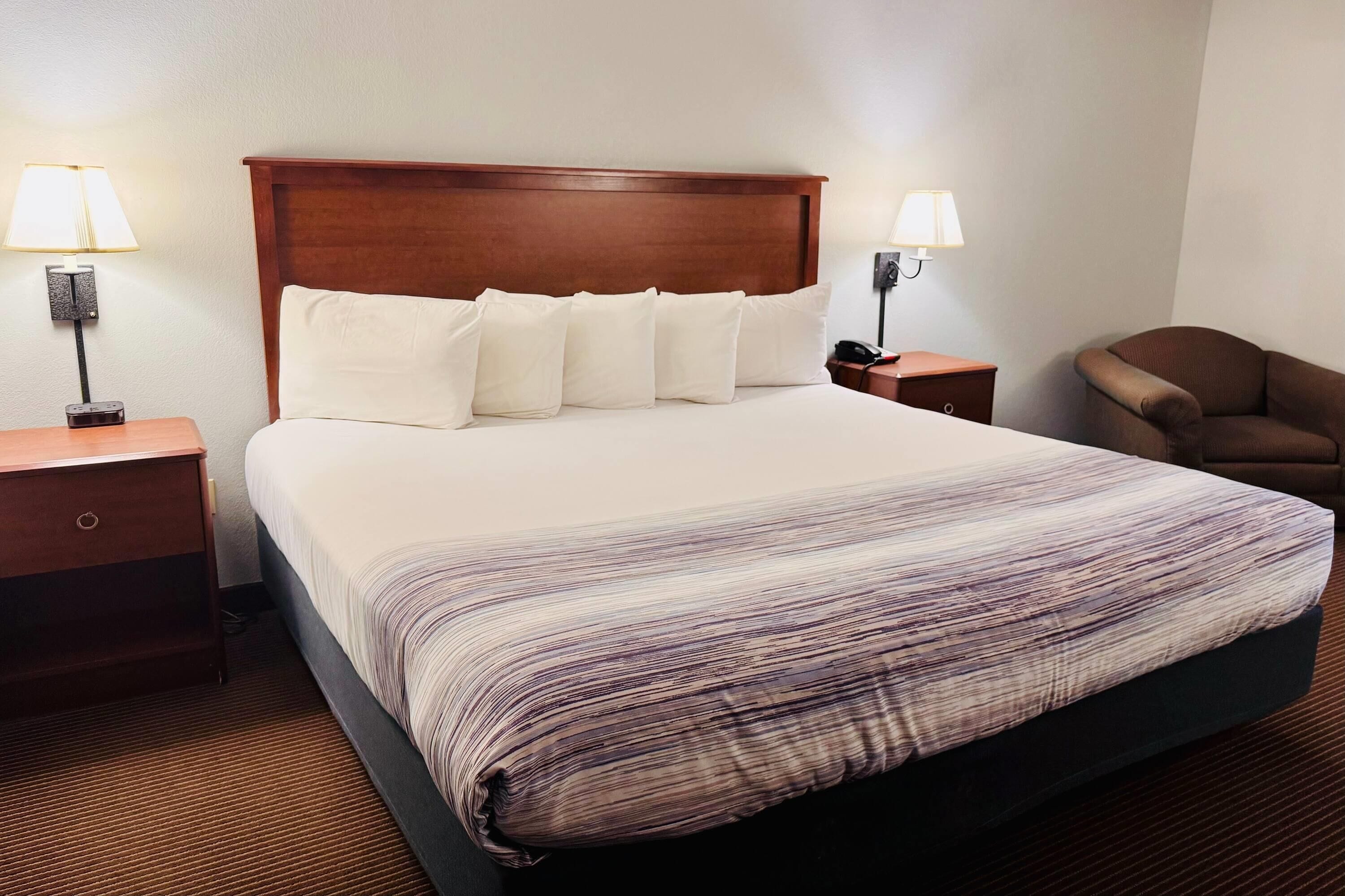 Quarto de hotel com uma cama grande, duas mesas de cabeceira com abajures, uma cadeira e uma cabeceira de madeira.