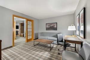 Quarto de hotel com sofá, mesa de centro, escrivaninha com luminária, pintura na parede e carpete xadrez.