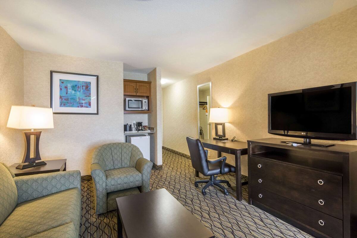 Quarto de hotel com sofá, poltrona, escrivaninha, televisão, cômoda, micro-ondas e uma moldura na parede.