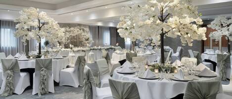 Uma sala de banquetes com mesas redondas preparadas para um evento formal, decorada com arranjos florais brancos e elegantes louças.
