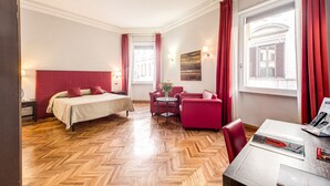 Quarto de hotel com cama, sofá vermelho, mesa pequena e escrivaninha com laptop e controle remoto.