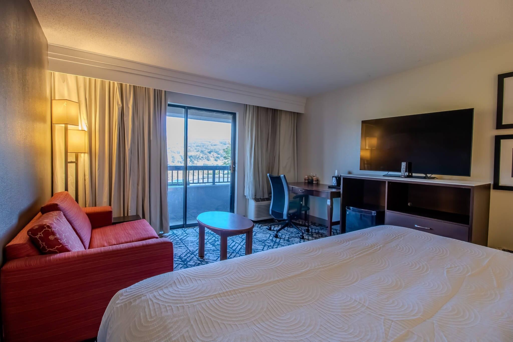 Quarto de hotel com uma cama grande, um sofá vermelho, uma escrivaninha com cadeira, televisão e vista para o exterior.