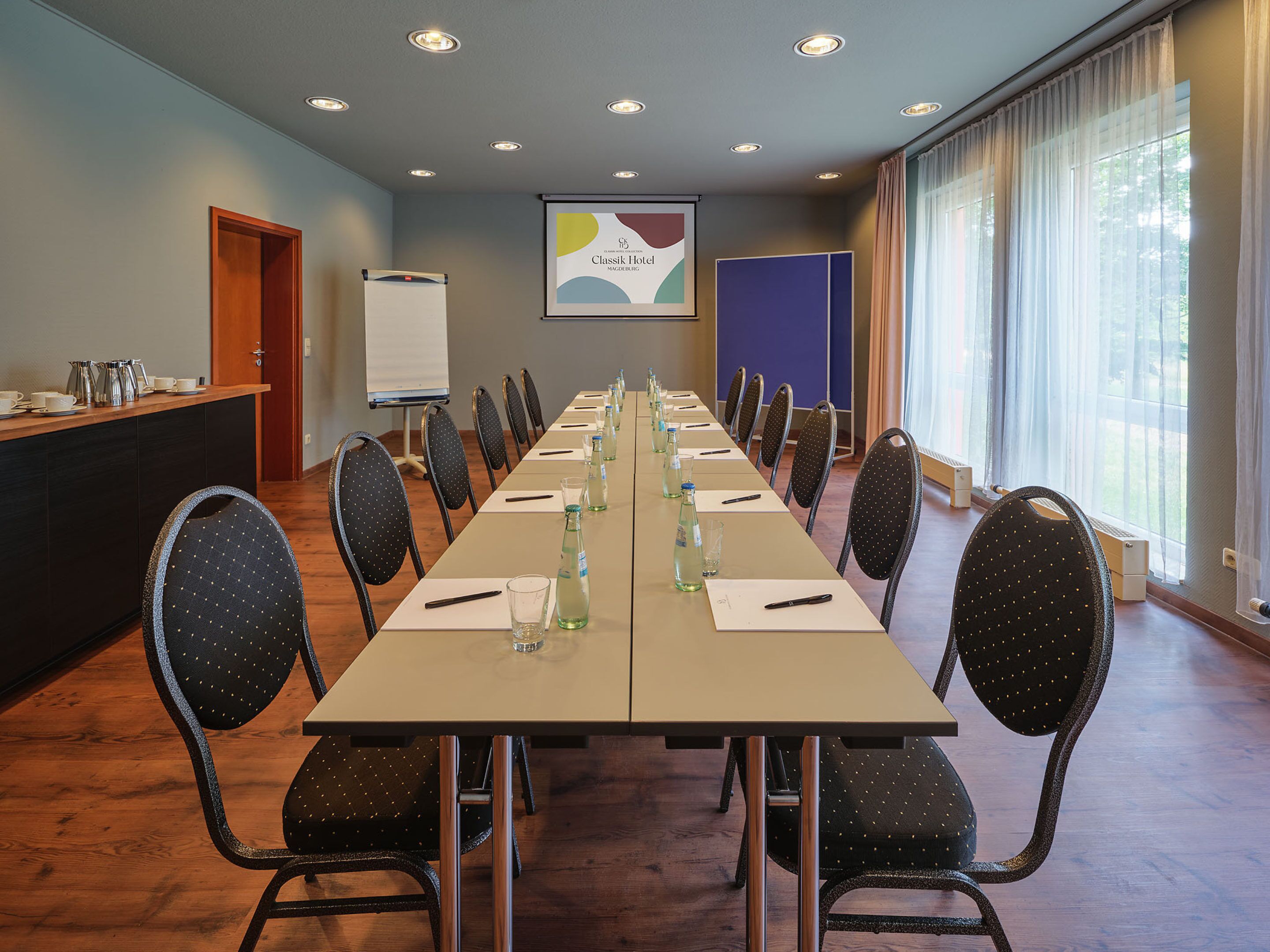 Sala de conferências com uma mesa comprida, cadeiras, tela de projetor e um canto para café.
