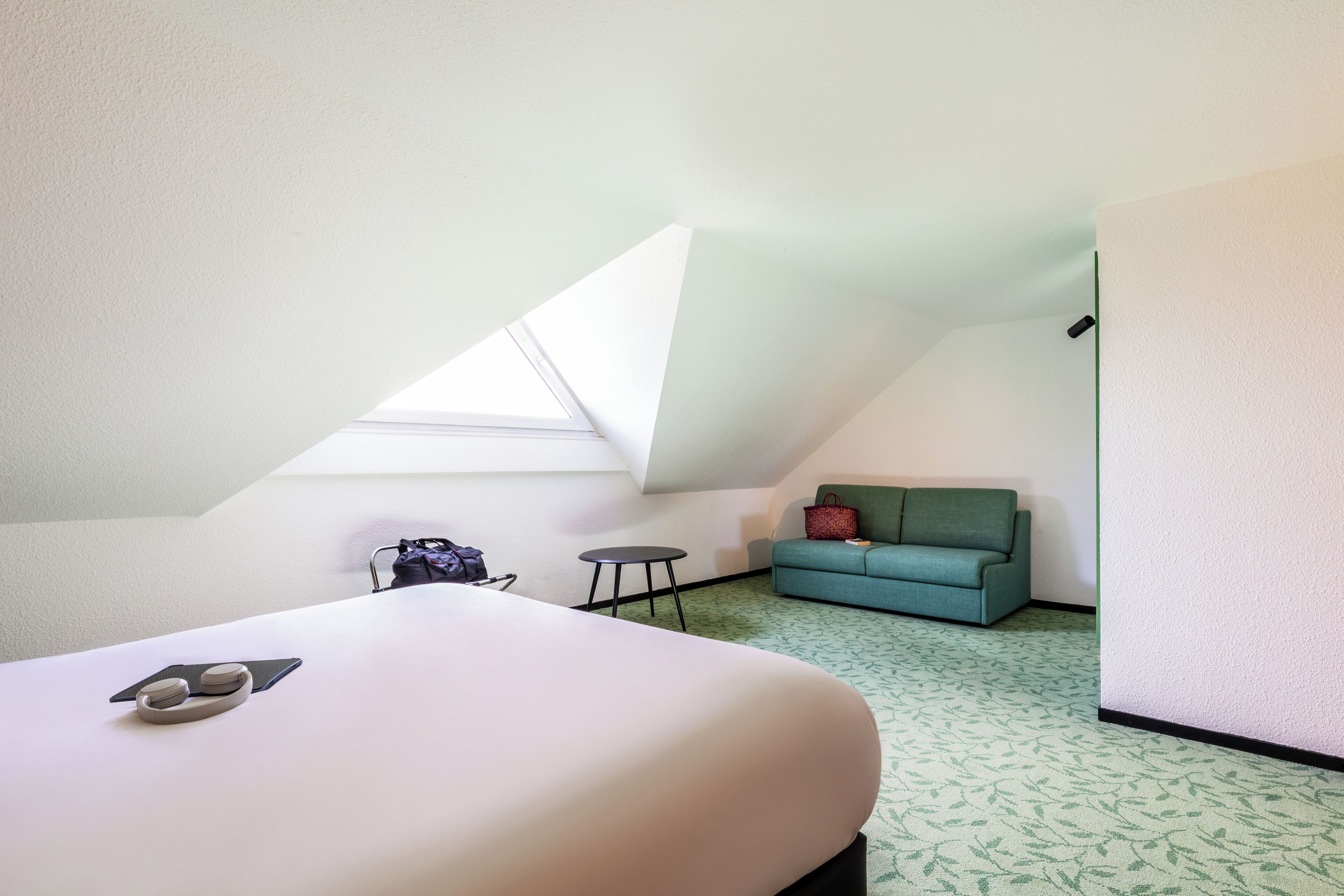 Quarto de hotel com cama, sofá verde, mesa pequena e janela.