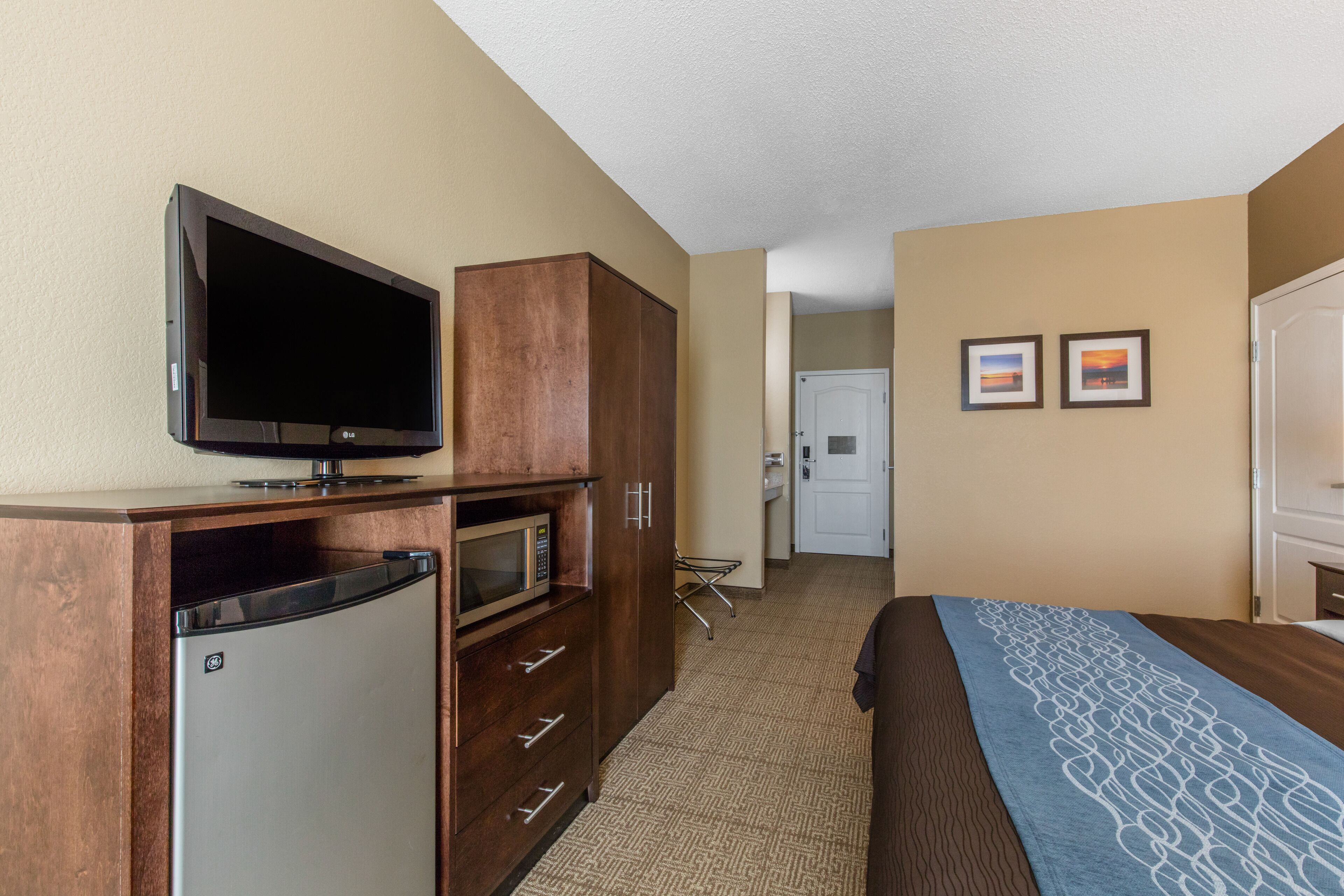 Quarto de hotel com cama, televisão, frigorífico e micro-ondas. Há uma porta que dá para uma casa de banho e um corredor.