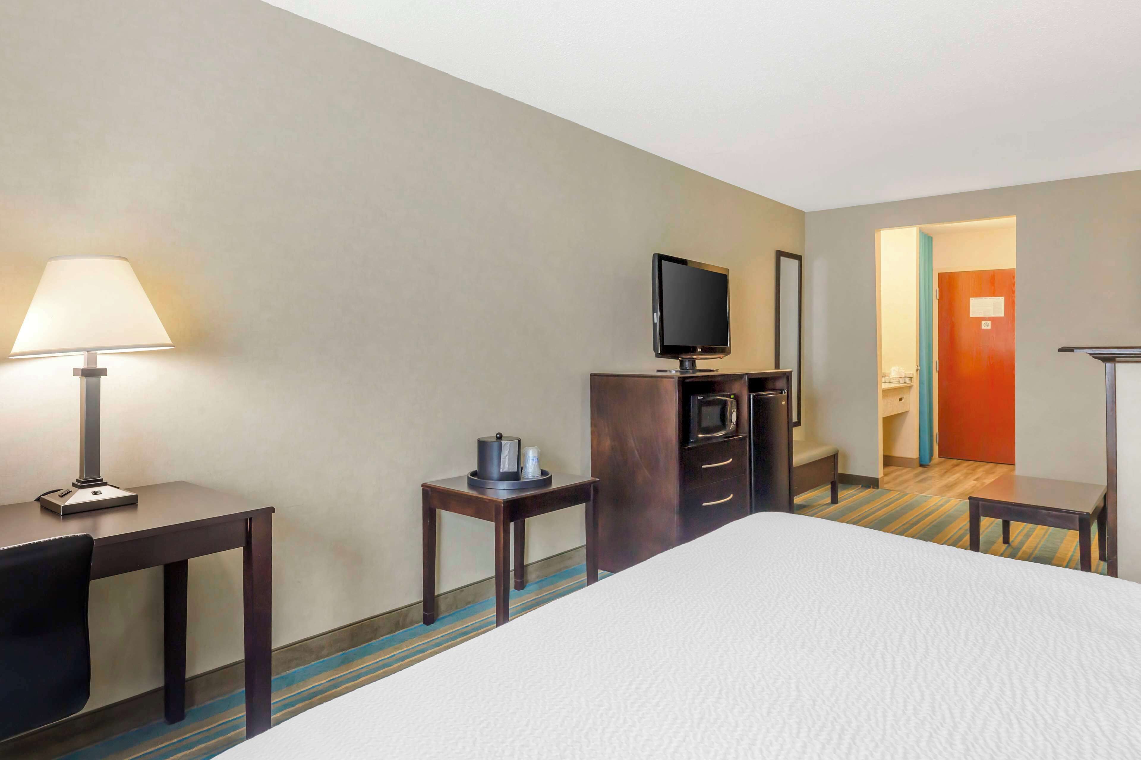 Quarto de hotel com cama, escrivaninha com luminária, televisão, micro-ondas e uma pequena mesa com uma xícara.