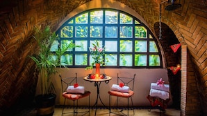 Um quarto aconchegante com lareira de tijolos, uma grande janela em arco, duas cadeiras, uma mesa com um candeeiro e um vaso de planta.