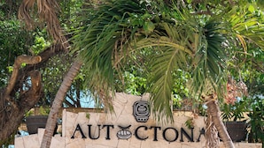 Entrada para um restaurante chamado "AUTCTONA", com arco de pedra, palmeiras e mesas ao ar livre.
