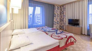 Quarto de hotel com cama, televisão sobre suporte, janela com cortinas e vista para prédios.