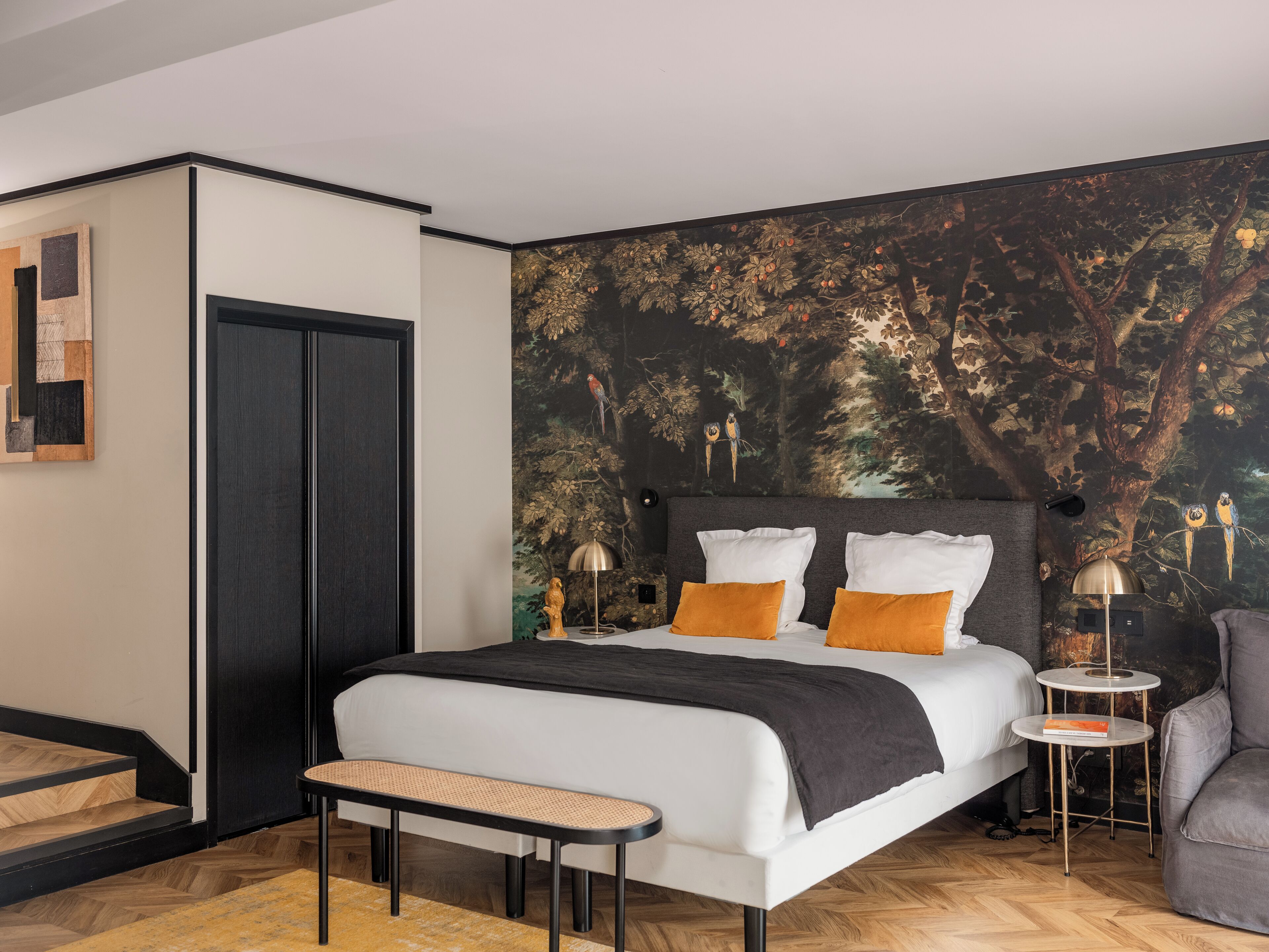 Quarto com cama, duas mesinhas de cabeceira, um banco, uma porta e parede com mural de árvore.