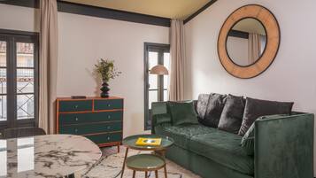 Uma sala de estar com um sofá verde, um espelho redondo, uma pequena mesa com um livro, um armário verde e um tapete estampado.