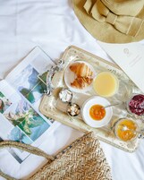 Uma bandeja com croissants, geleias e um copo de suco sobre uma mesa com uma revista e uma bolsa de palha.