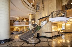 Um amplo saguão de hotel com escadaria central, vários níveis e uma escultura de metal decorativa.