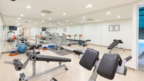 Uma academia com diversos equipamentos de exercício, um espelho grande e uma televisão na parede.