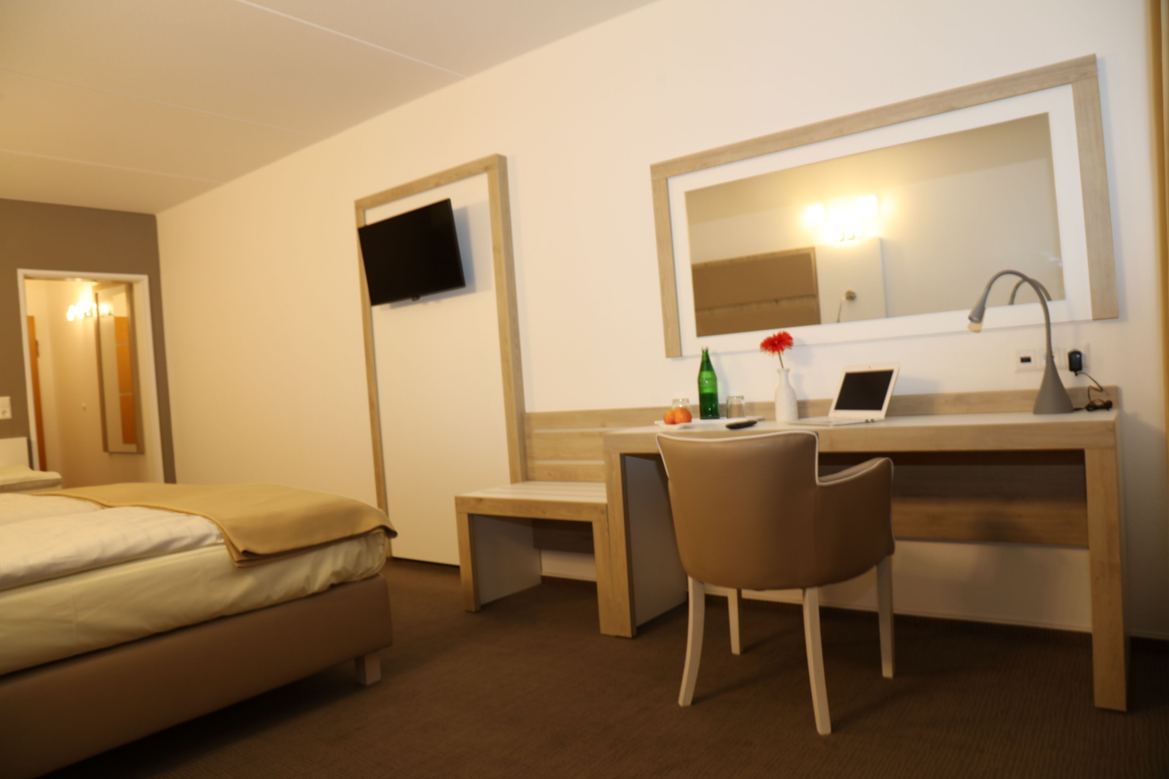 Quarto de hotel com cama, escrivaninha, cadeira, laptop, espelho e televisão.