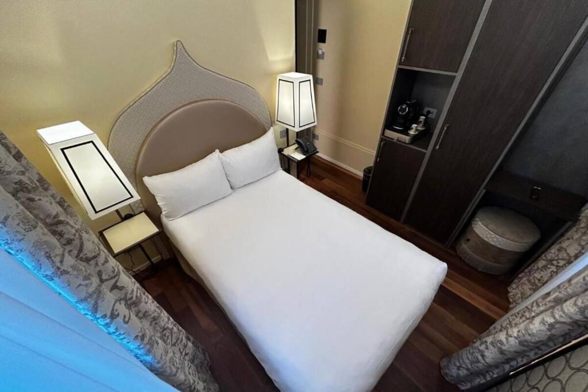 Cama bem arrumada com lençóis brancos, mesa de cabeceira com abajur e guarda-roupa em um quarto de hotel.