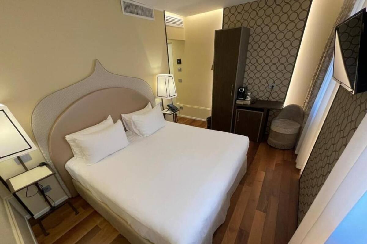 Quarto de hotel com uma cama grande, mesa de cabeceira, luminária, guarda-roupa e um espelho.