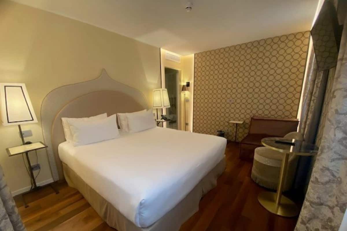 Quarto de hotel com cama grande, abajures, uma cadeira e uma mesinha.