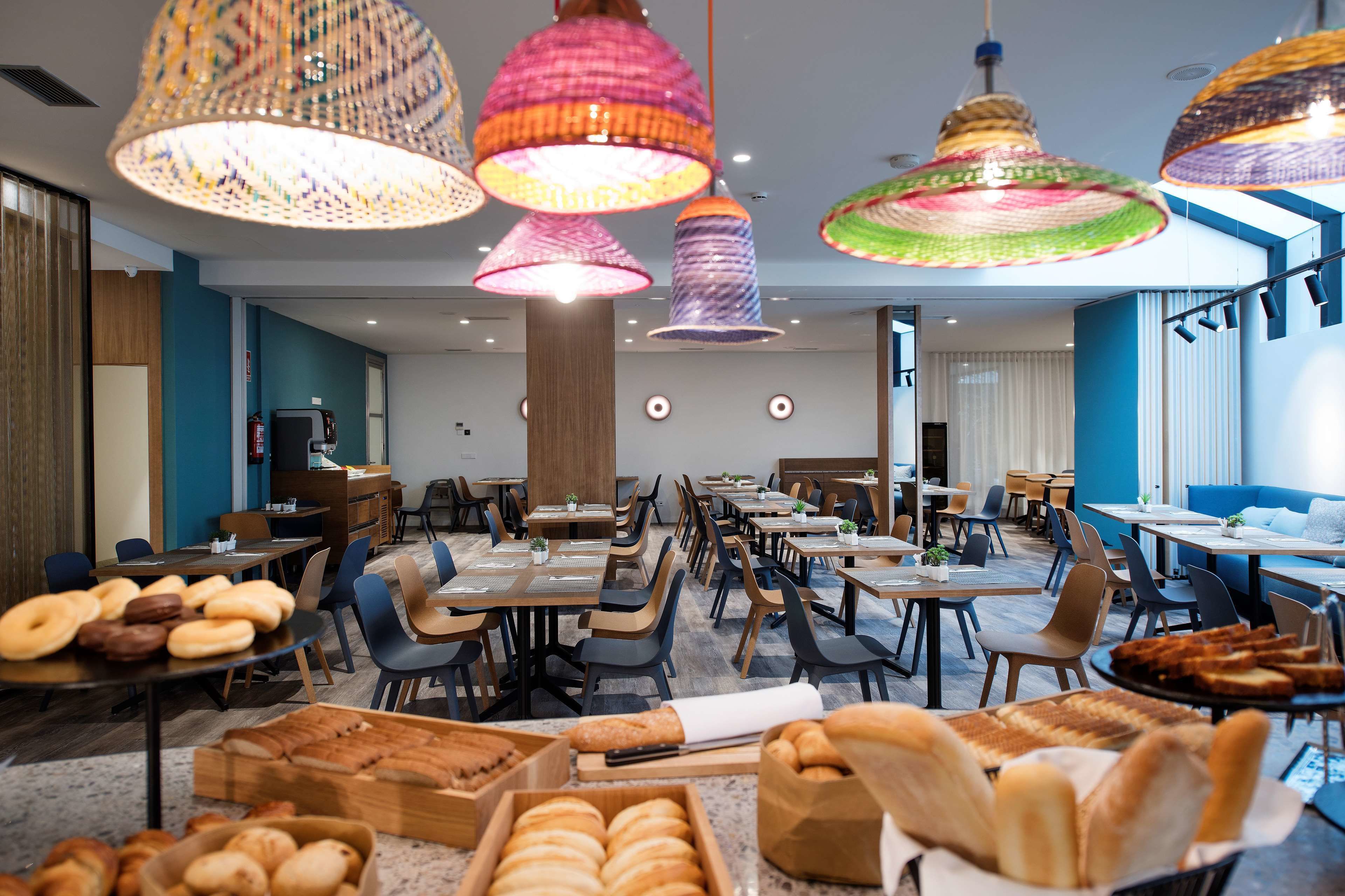 Uma sala de jantar moderna com várias mesas e cadeiras, luminárias pendentes coloridas e uma variedade de pães expostos sobre as mesas.