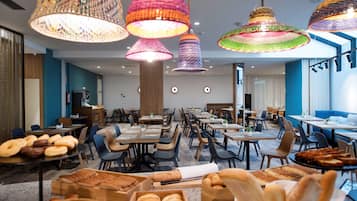 Uma sala de jantar moderna com várias mesas e cadeiras, luminárias pendentes coloridas e uma variedade de pães expostos sobre as mesas.
