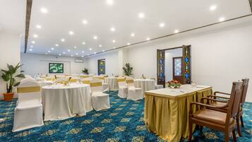 Sala de conferência com mesas redondas, cadeiras brancas com fitas douradas e um tapete com padrão azul.