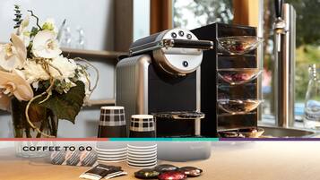 Uma máquina de café Nespresso com vários pões de café e um jarro, um vaso com flores brancas e uma janela com vista para vegetação.
