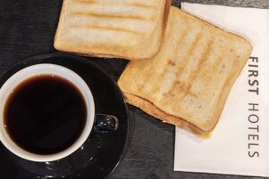 Sanduíche e uma chávena de café sobre uma mesa preta, com uma guardanapo e uma placa a indicar "FIRST HOTELS".