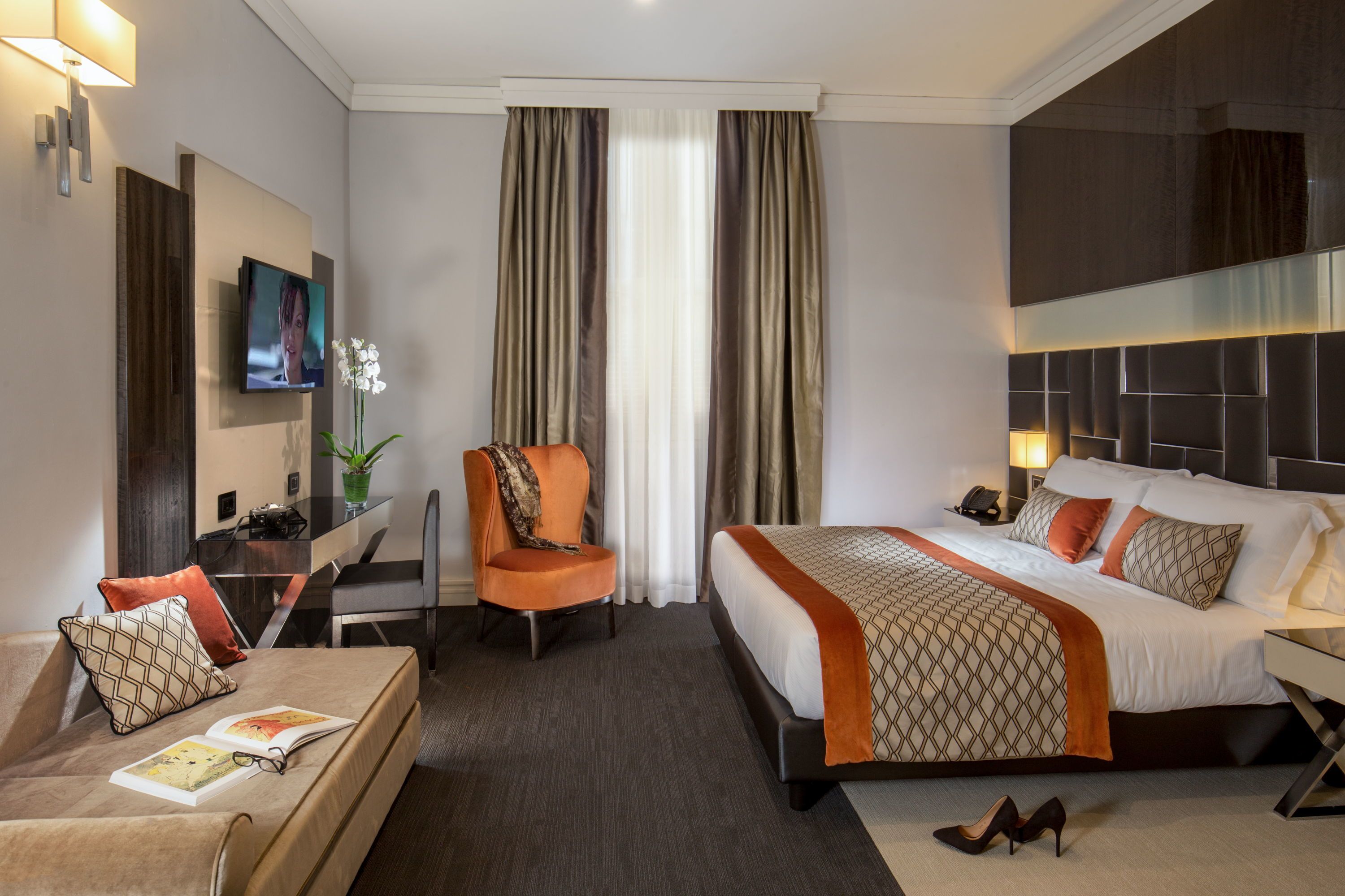 Quarto de hotel com cama, escrivaninha, cadeira, televisão e janela com cortinas.