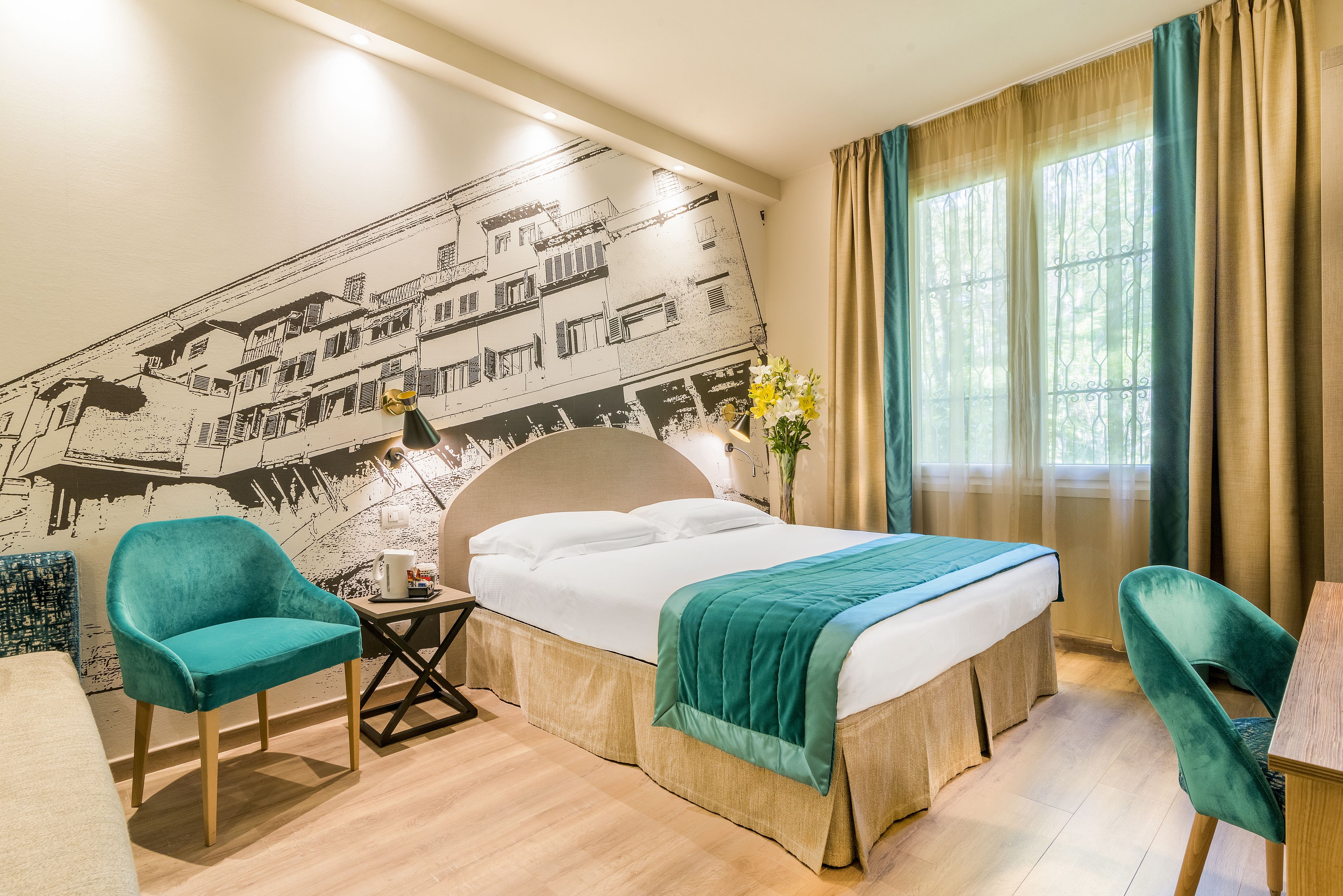 Quarto de hotel com cama, duas cadeiras, uma mesa pequena com abajur, um vaso com flores e um mural grande na parede.