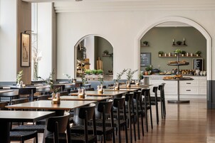 Um restaurante com várias mesas e cadeiras, uma área de cozinha, e prateleiras com plantas e alimentos.