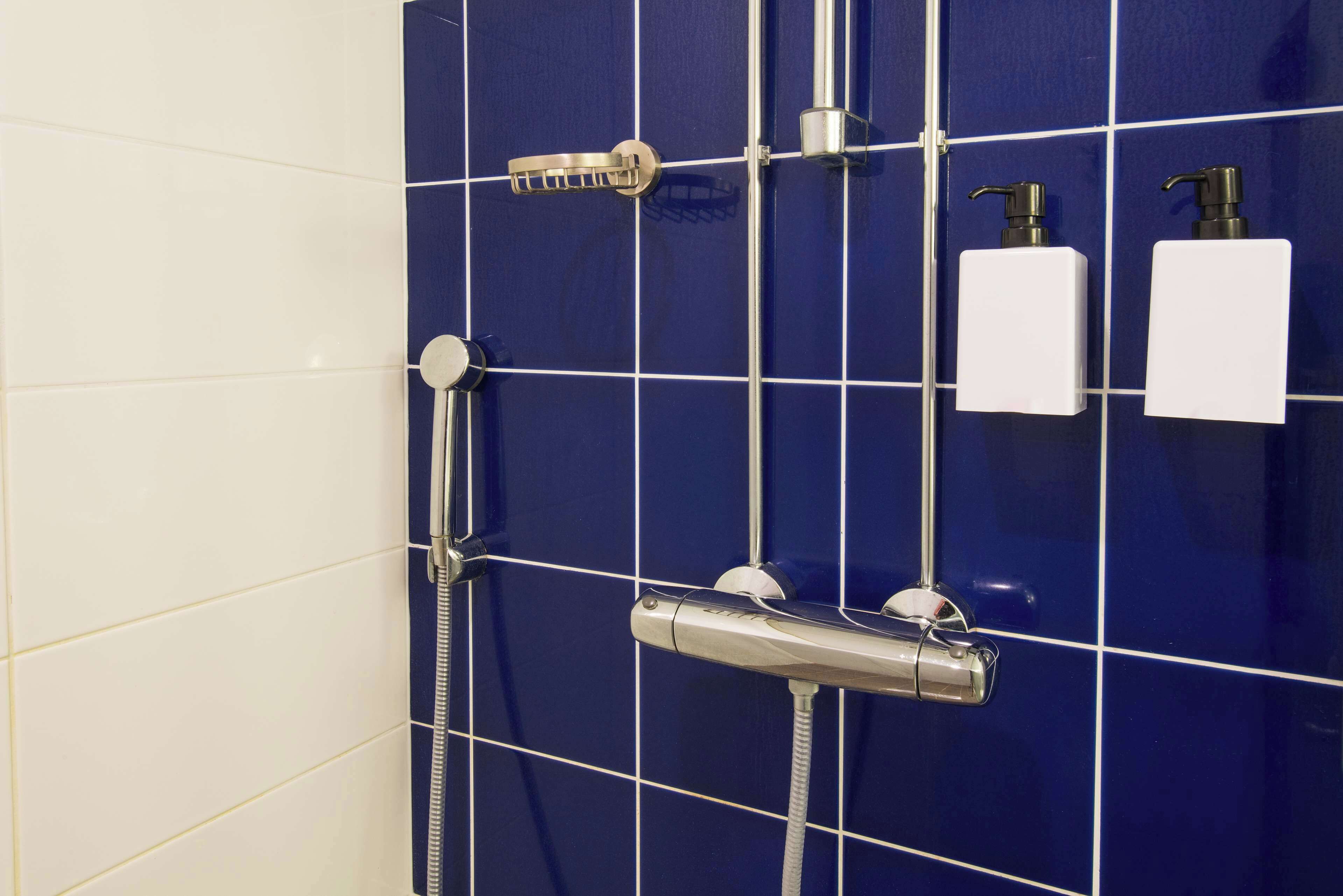 Casa de banho com parede revestida em azulejos azuis, chuveiro fixo e chuveiro manual, e dois dispensadores brancos.