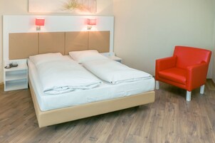 Uma cama com duas almofadas, uma mesa de cabeceira, uma cadeira vermelha e duas luminárias na parede.