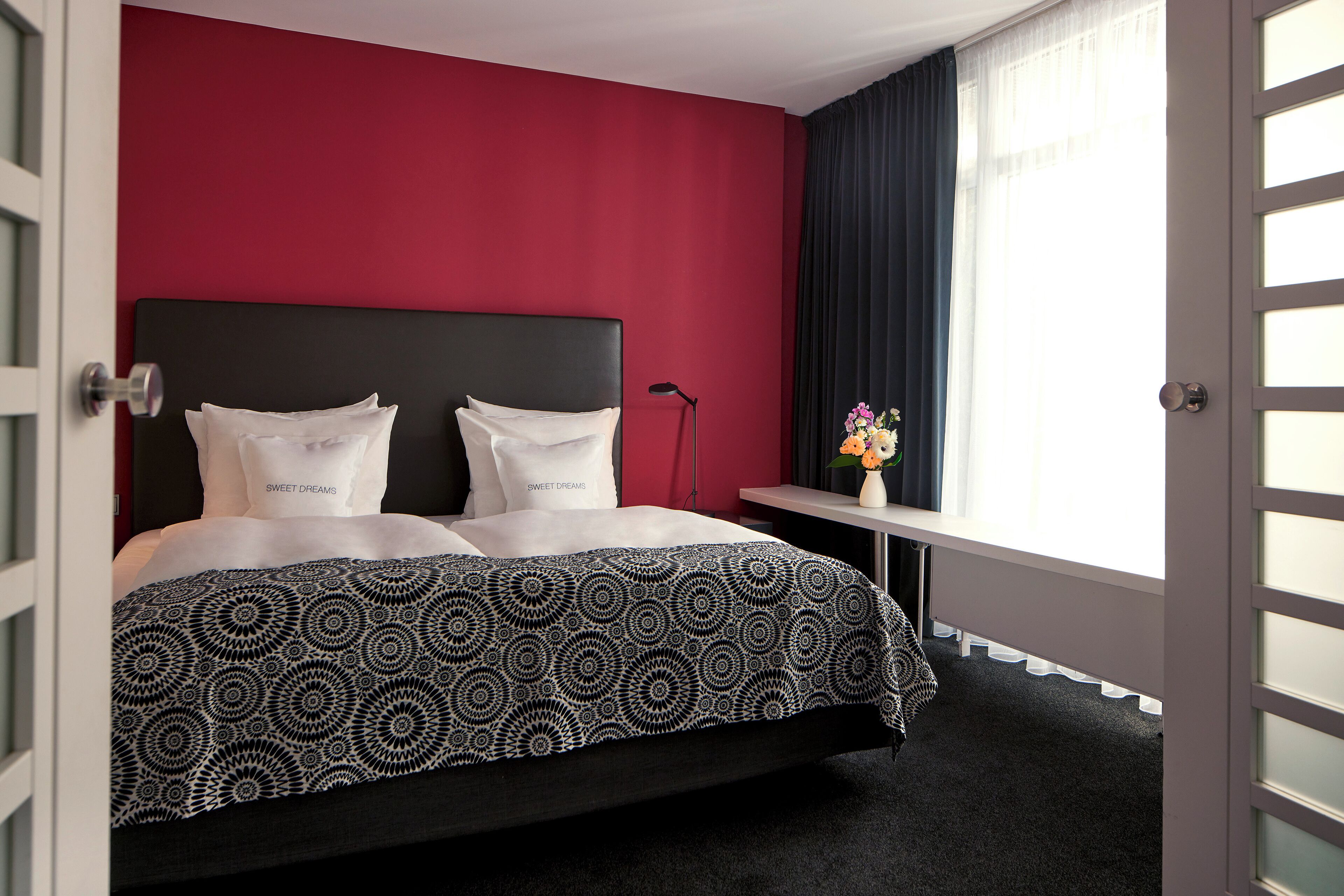 Quarto de hotel com parede vermelha, cabeceira preta, colcha estampada, mesa de cabeceira com um vaso de flores e janela com cortinas.