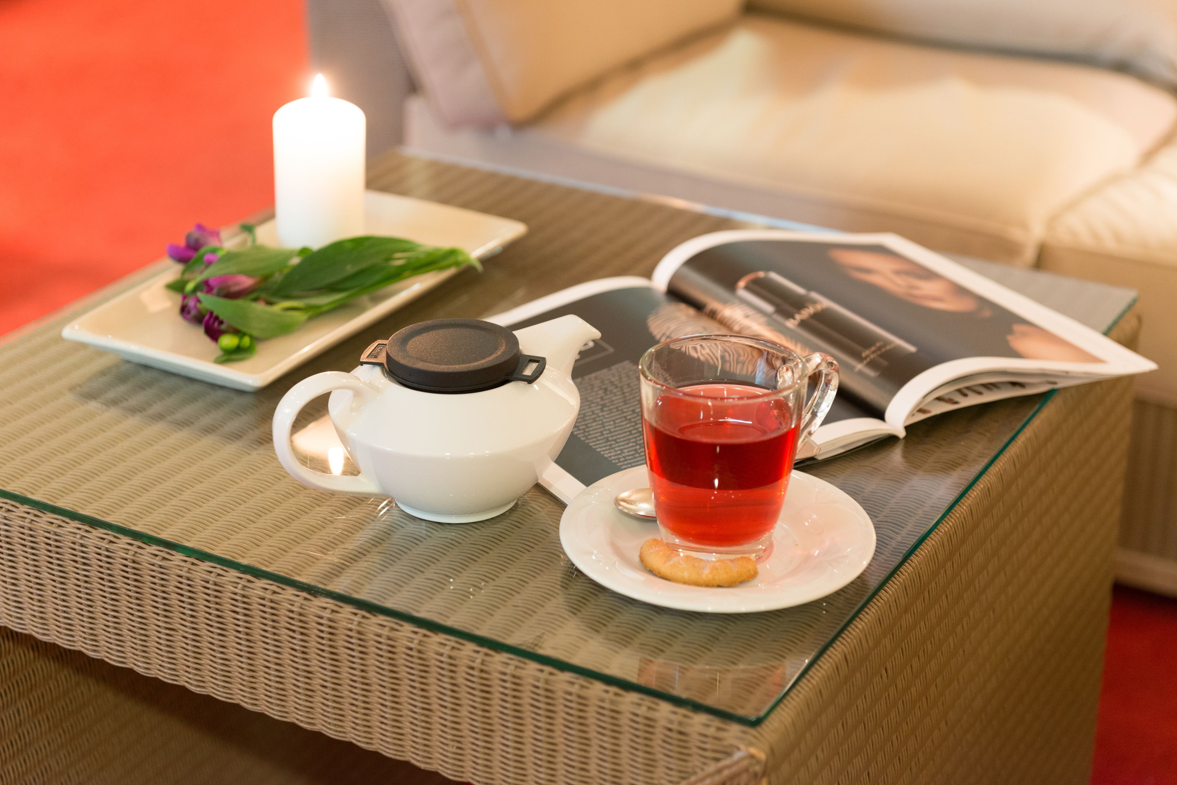Uma mesa de vidro com uma chaleira, uma chávena de chá, um prato com um biscoito, uma vela e uma revista.