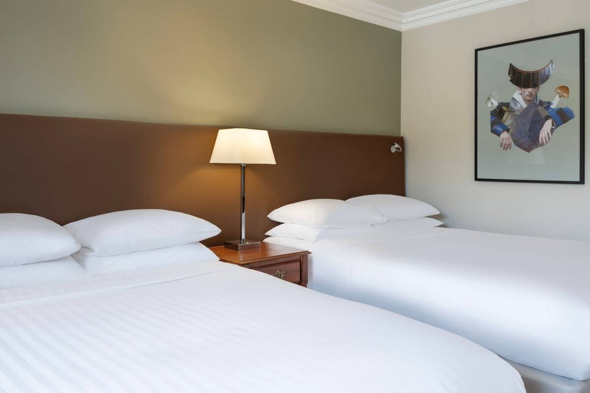 Quarto de hotel com duas camas, uma mesa de cabeceira com abajur e uma obra de arte emoldurada na parede.