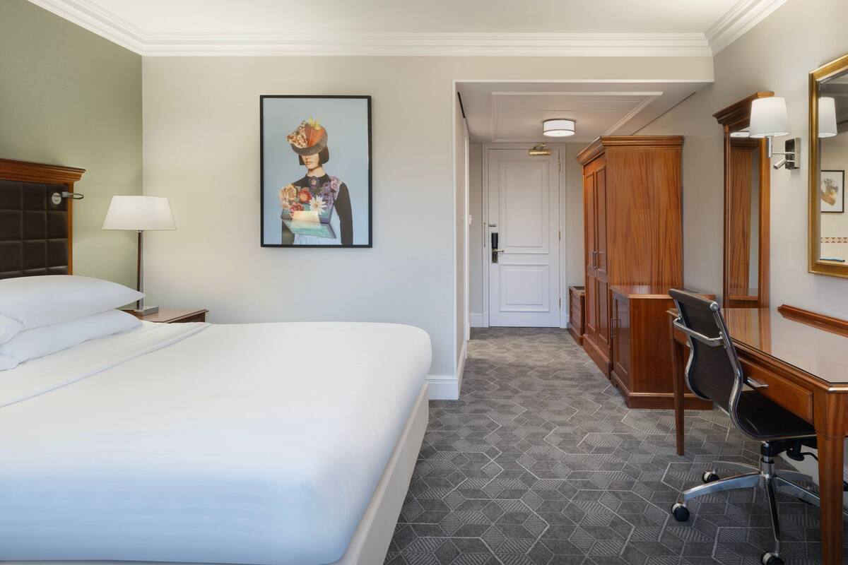 Quarto de hotel com uma cama grande, uma escrivaninha com cadeira, um guarda-roupa, um quadro na parede e um tapete estampado.