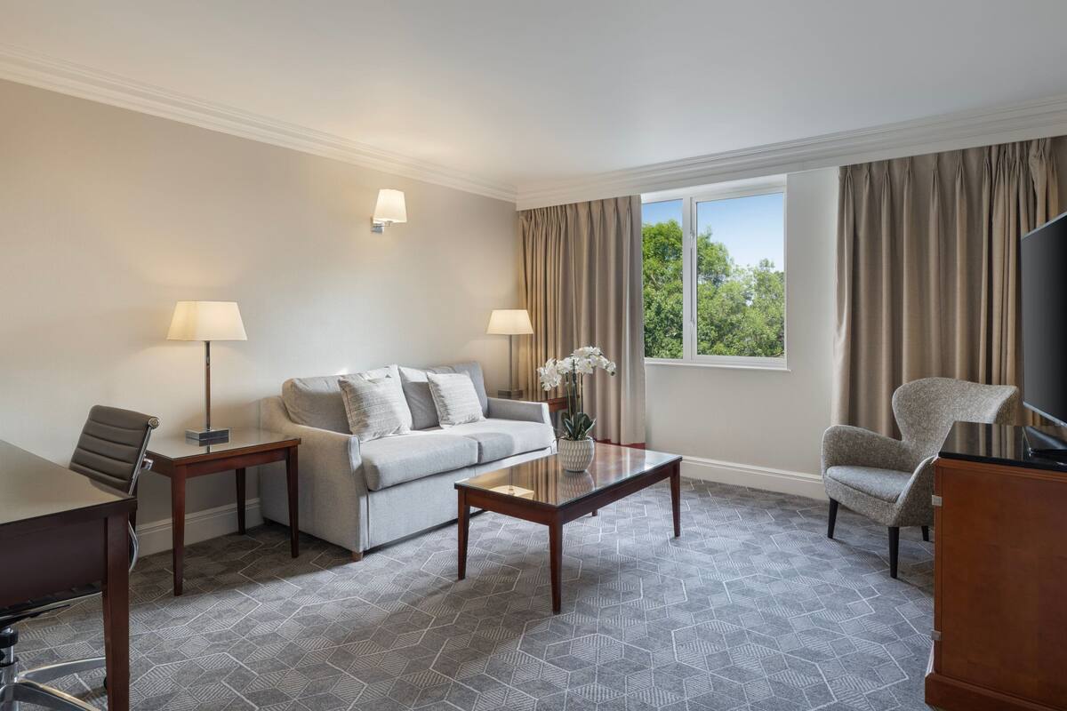 Quarto de hotel com sofá, duas poltronas, mesa de centro, televisão e janela com cortinas.