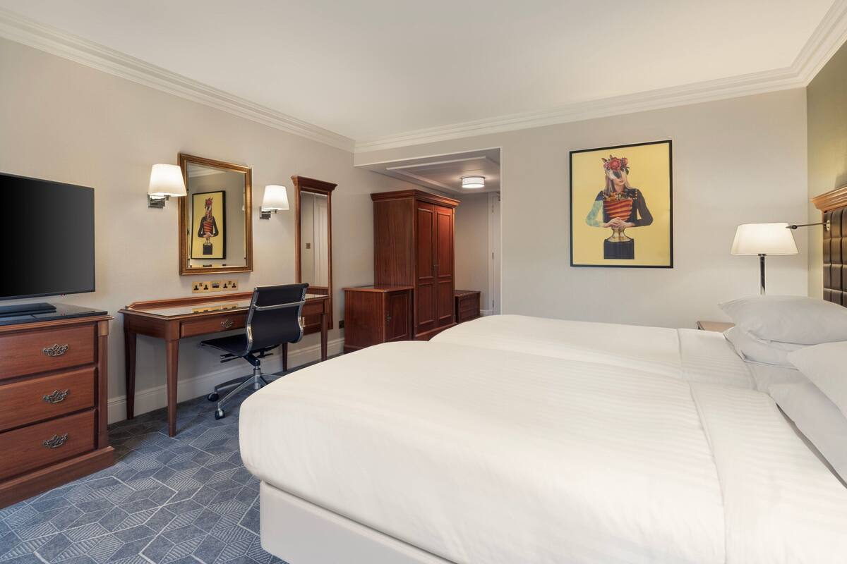 Quarto de hotel com uma cama grande, uma escrivaninha com computador, uma cadeira, televisão, guarda-roupa e um quadro na parede.