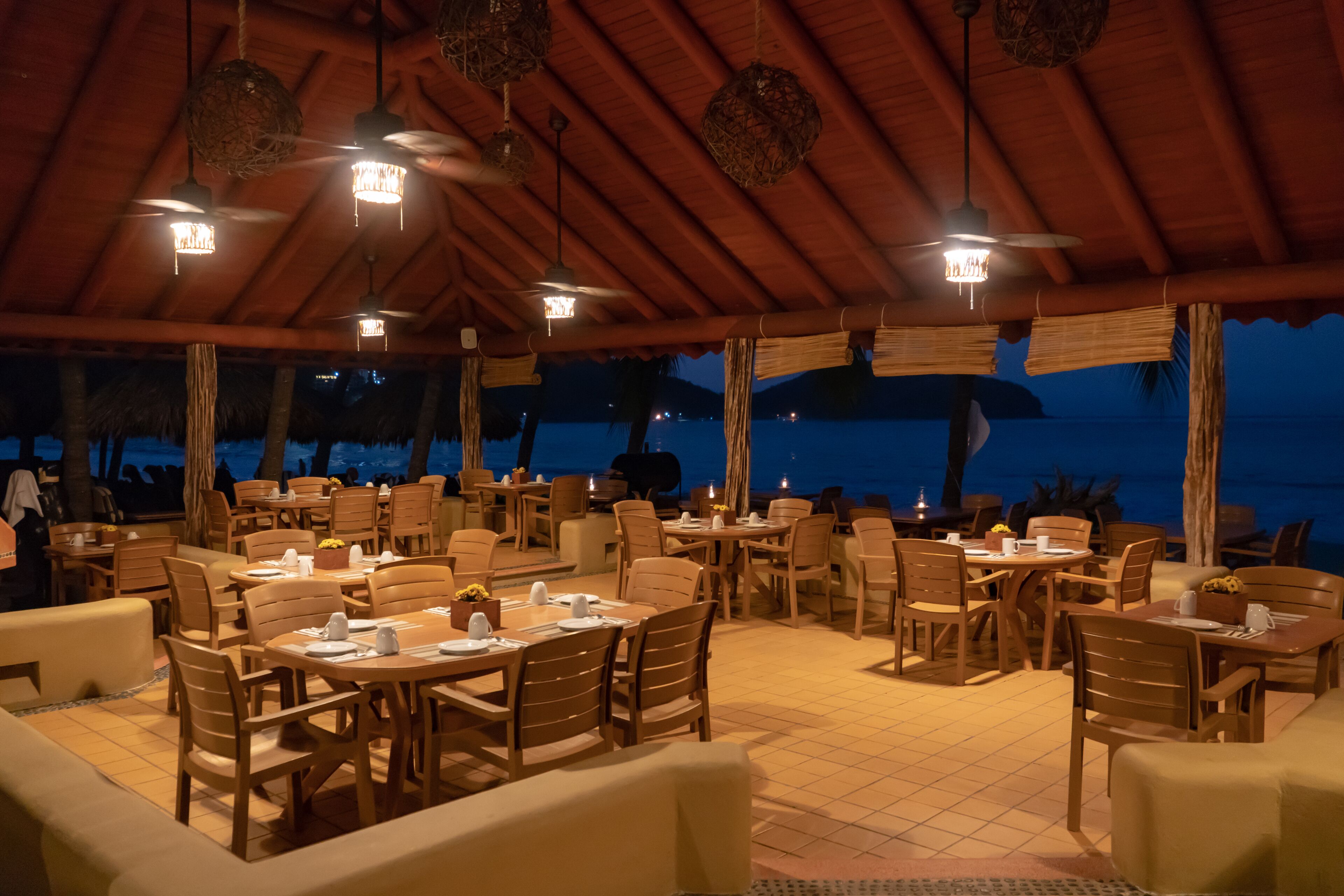 Uma área de refeições ao ar livre com mesas e cadeiras de madeira, luminárias pendentes e vista para o oceano ao entardecer.