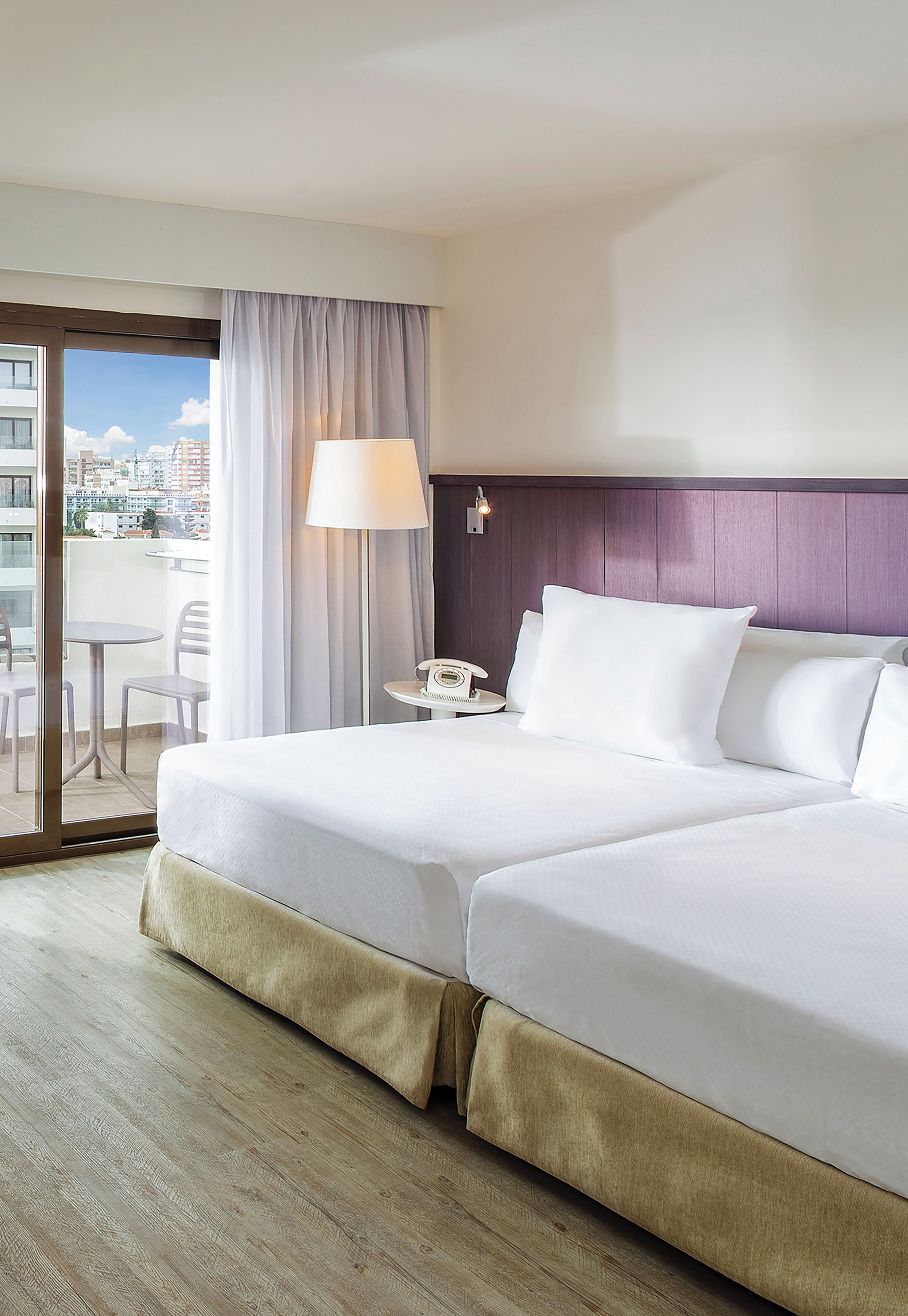 Quarto de hotel com uma cama grande, uma mesa de cabeceira com luminária e vista da cidade por uma porta de correr.