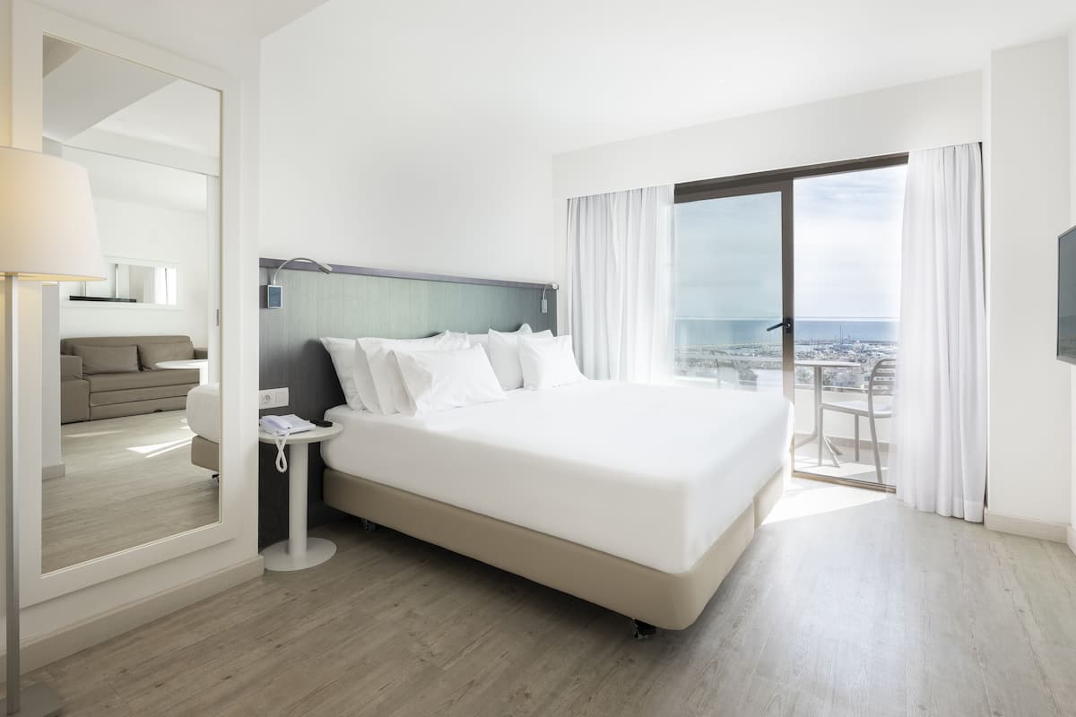 Quarto de hotel com cama, mesa de cabeceira, luminária, espelho, vista da cidade e varanda com cortinas.